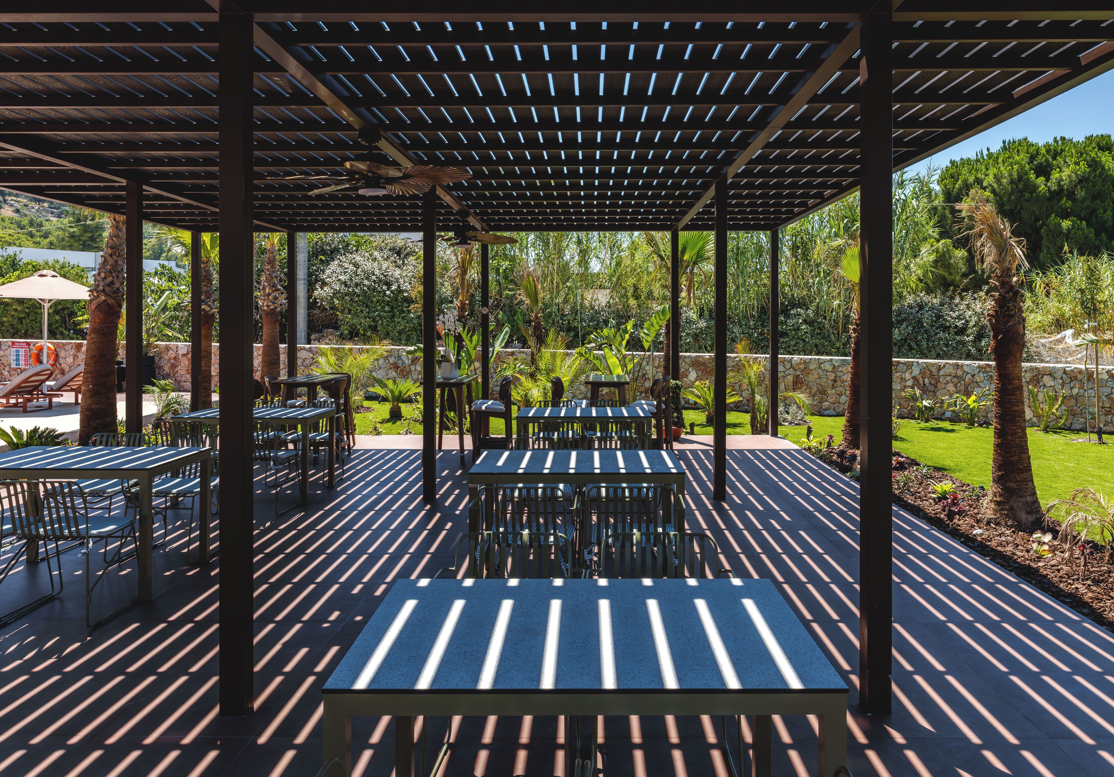 Um pátio ao ar livre com mesas e cadeiras, uma estrutura de pérgola e vista para áreas verdes e uma piscina.