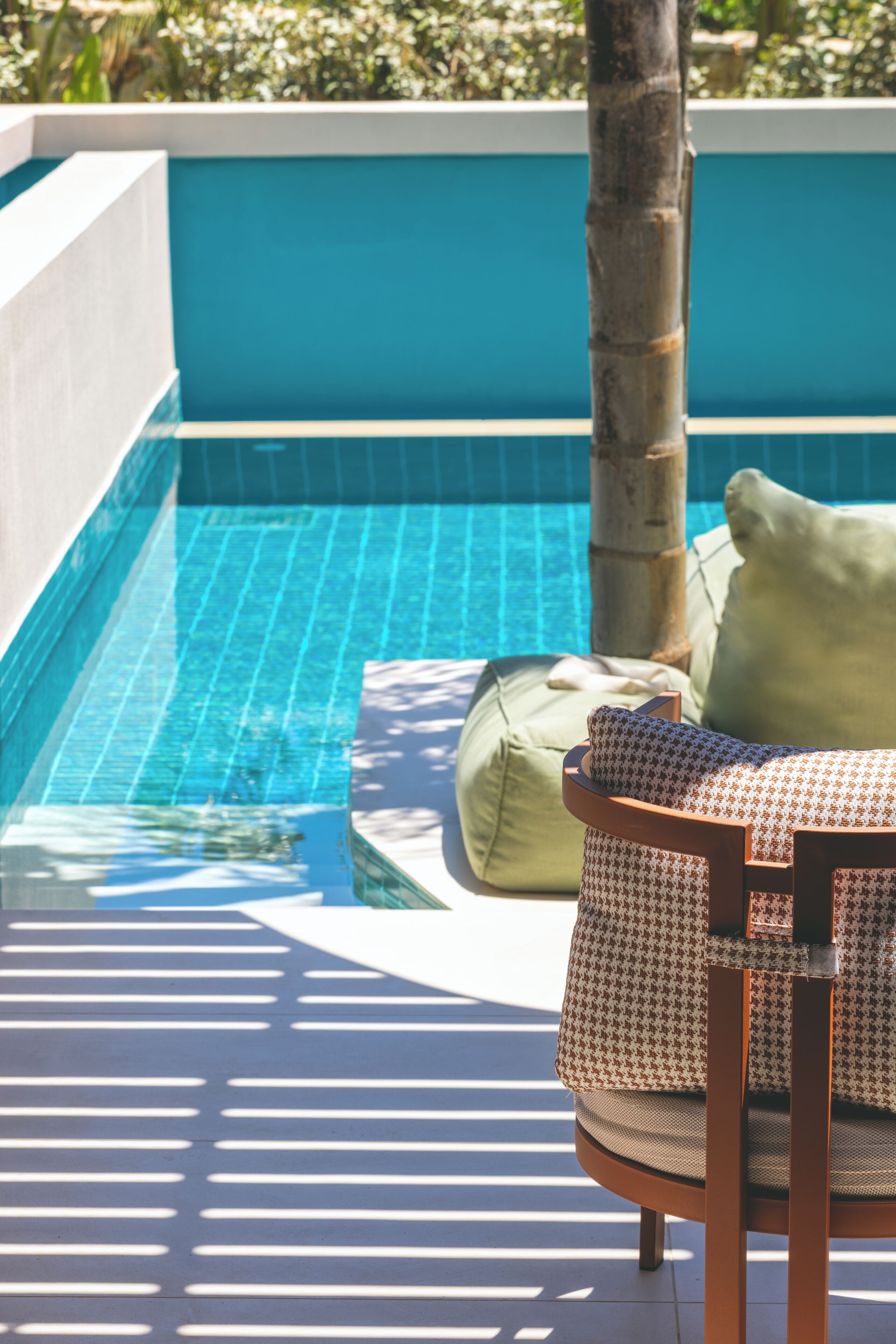 Uma área junto à piscina com uma cadeira, um almofada, uma árvore e uma piscina revestida com azulejos.