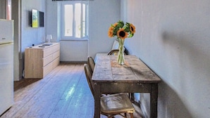 Quarto com uma mesa de madeira, um vaso com girassóis, uma televisão na parede e uma janela com persianas.