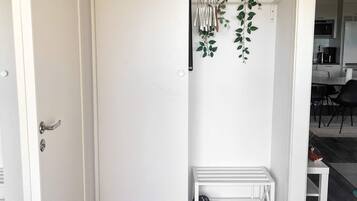 Uma porta branca com maçaneta, um banco branco com um objeto dobrado e uma planta fixada na parede.