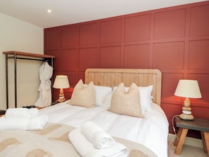 Quarto de hotel com cabeceira de madeira, roupa de cama branca, almofadas estampadas, mesa de cabeceira com abajur e roupão pendurado em um cabide.