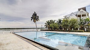 Piscina com vista clara para o mar, cercada por palmeiras e um edifício com varanda.