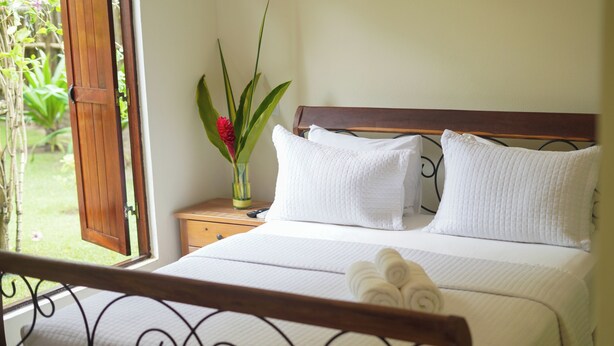 Cama bem arrumada com roupa de cama e almofadas brancas, mesa de cabeceira com um vaso de flores e janela com vista para vegetação.