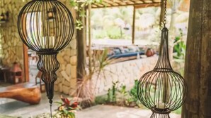 Uma área externa rústica com lanternas de metal penduradas, paredes de pedra e vegetação.