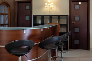 Uma área de recepção com balcão de vidro, banquetas pretas, um candelabro e portas de madeira com painéis de vidro jateado.