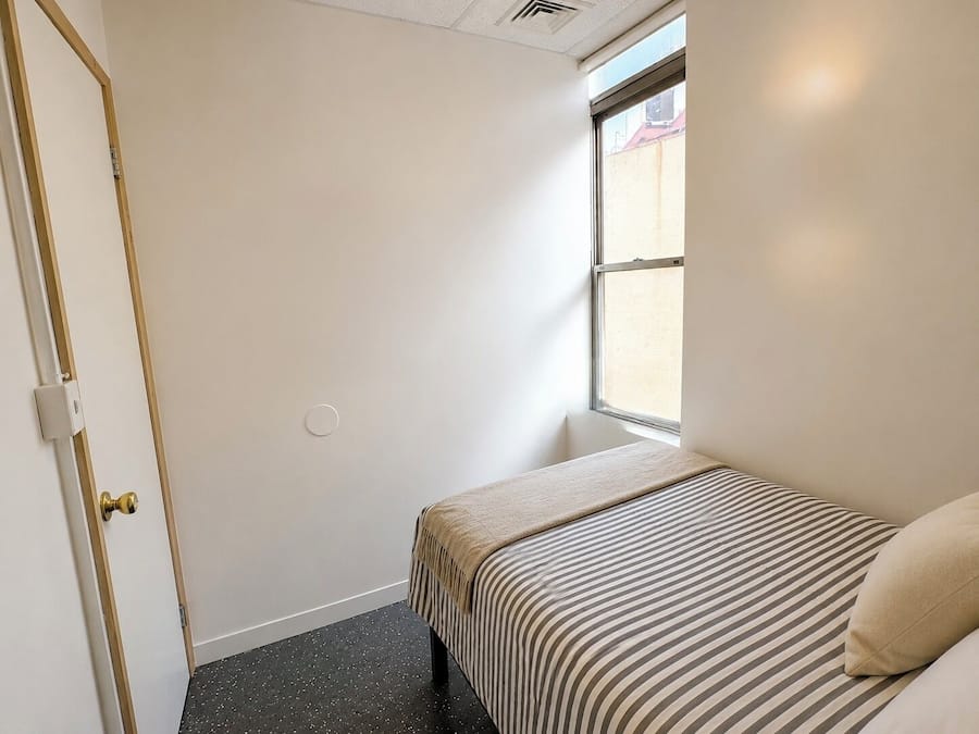 Quarto pequeno com cama de solteiro, janela, porta e chão coberto por carpete.