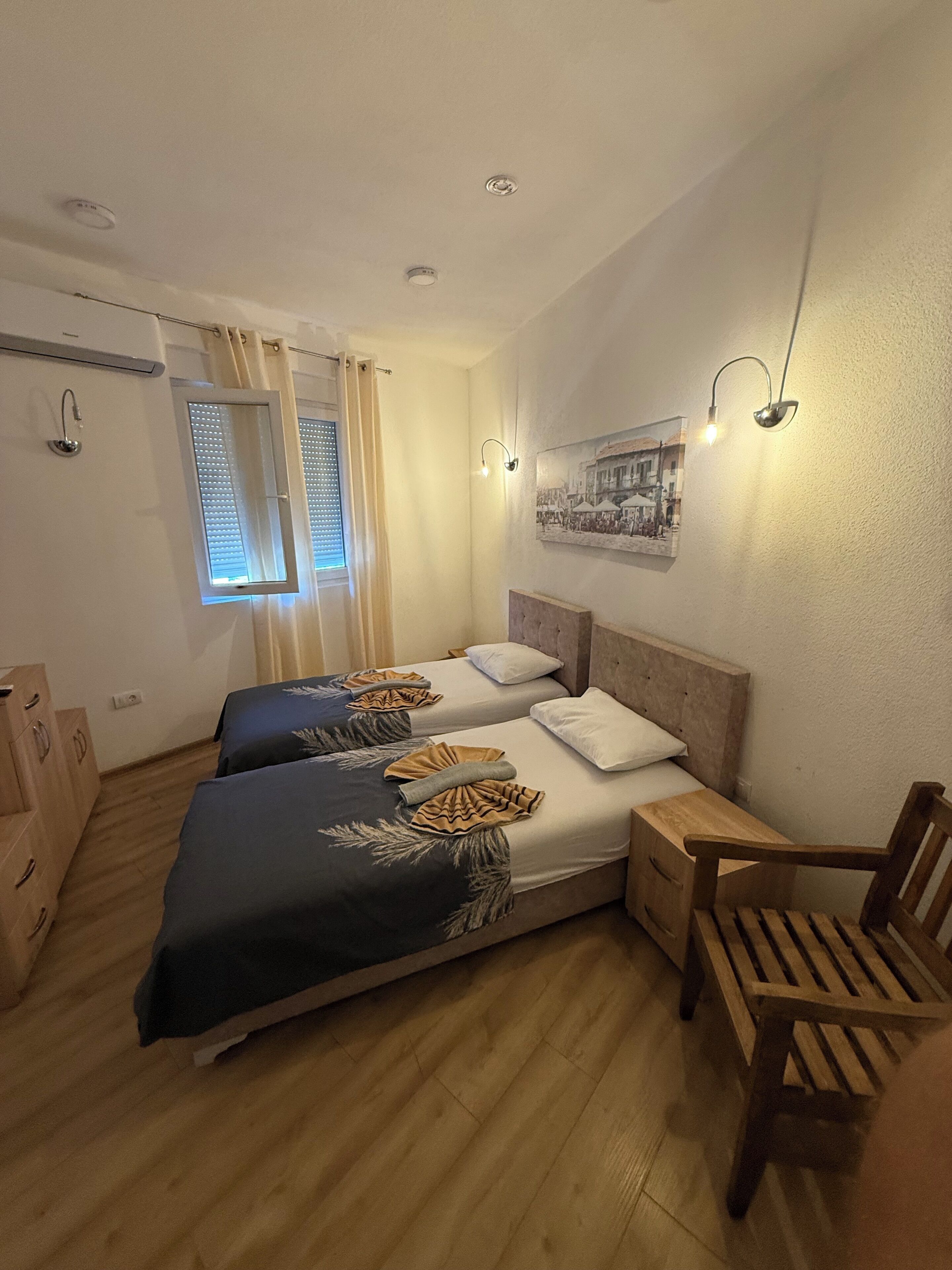 Quarto de hotel com duas camas, uma cadeira de madeira, uma mesinha, uma janela com persianas e um quadro pendurado na parede.