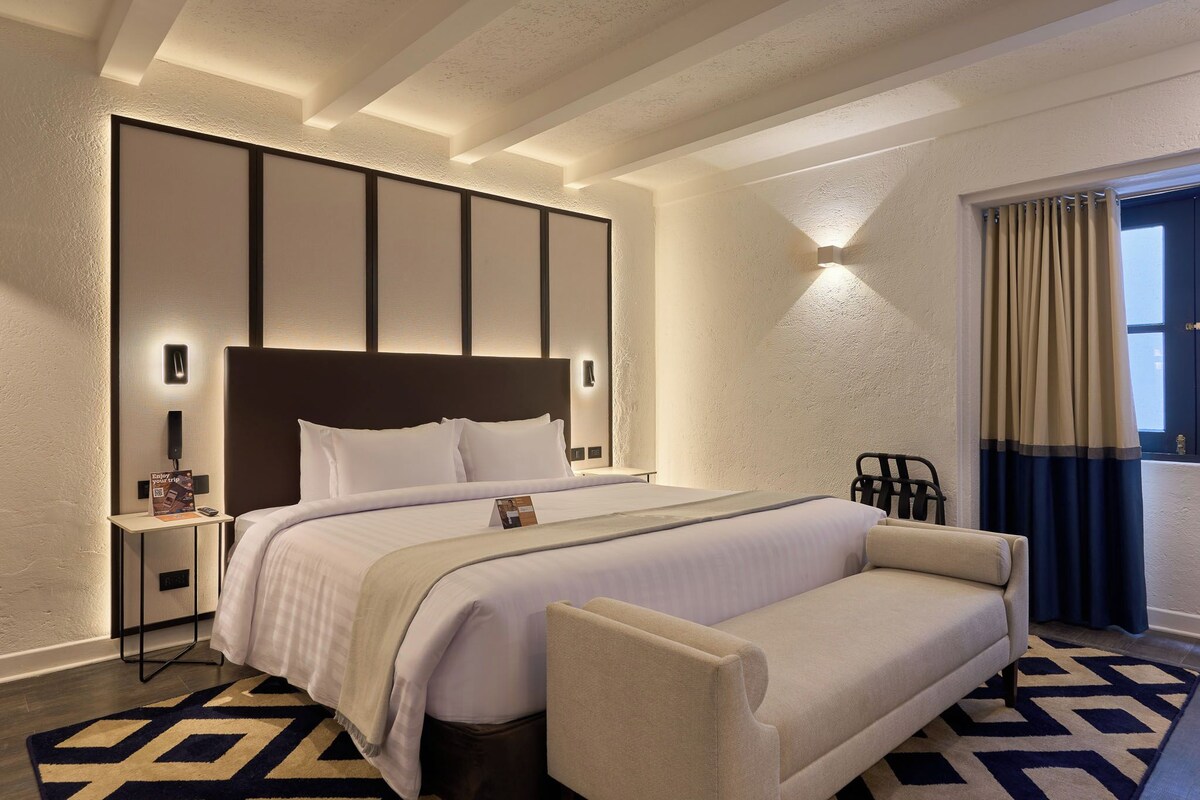 Quarto de hotel com uma cama grande, mesas de cabeceira, luminária na parede e vista para o exterior.