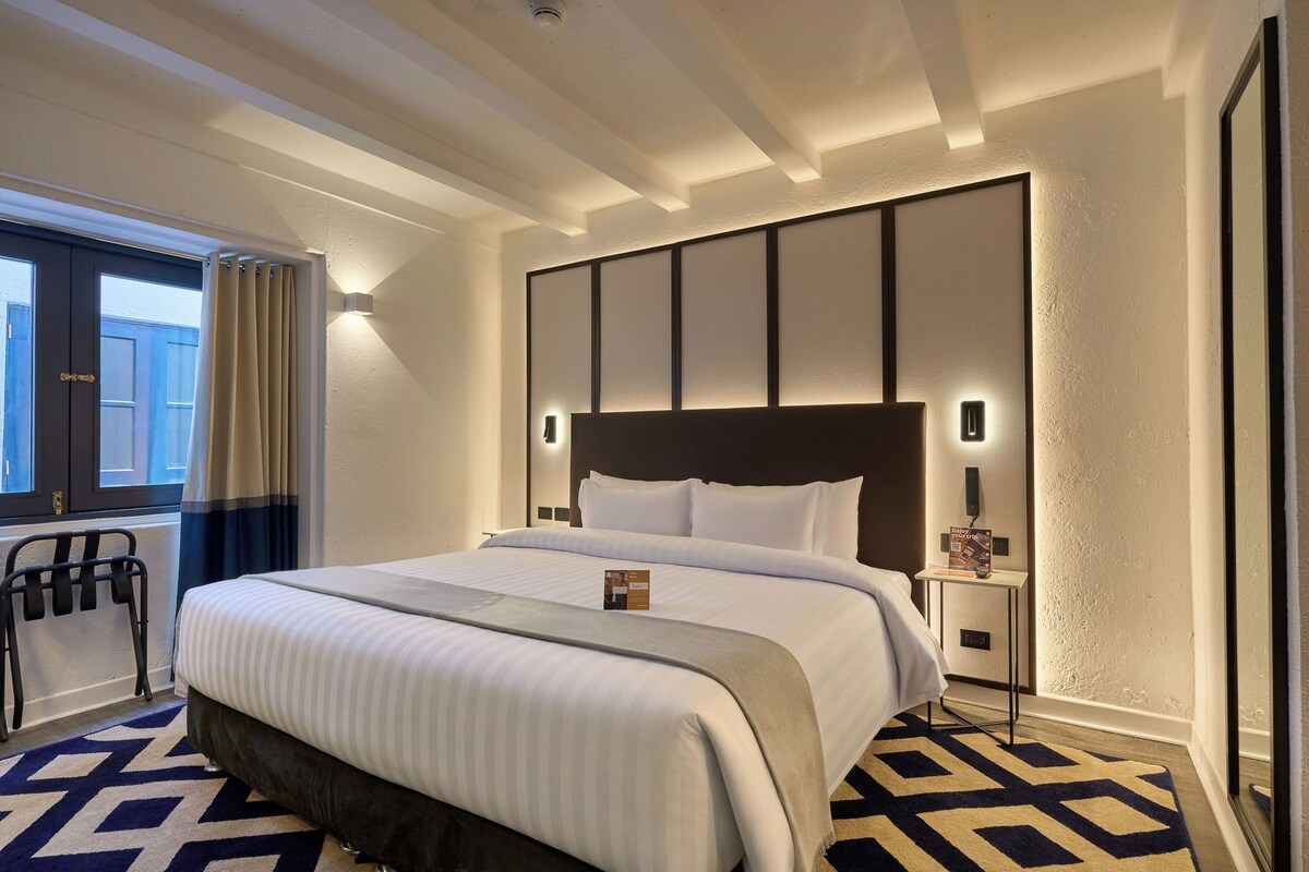 Quarto de hotel com uma cama grande, mesa de cabeceira com abajur, uma cadeira e vista para o exterior.