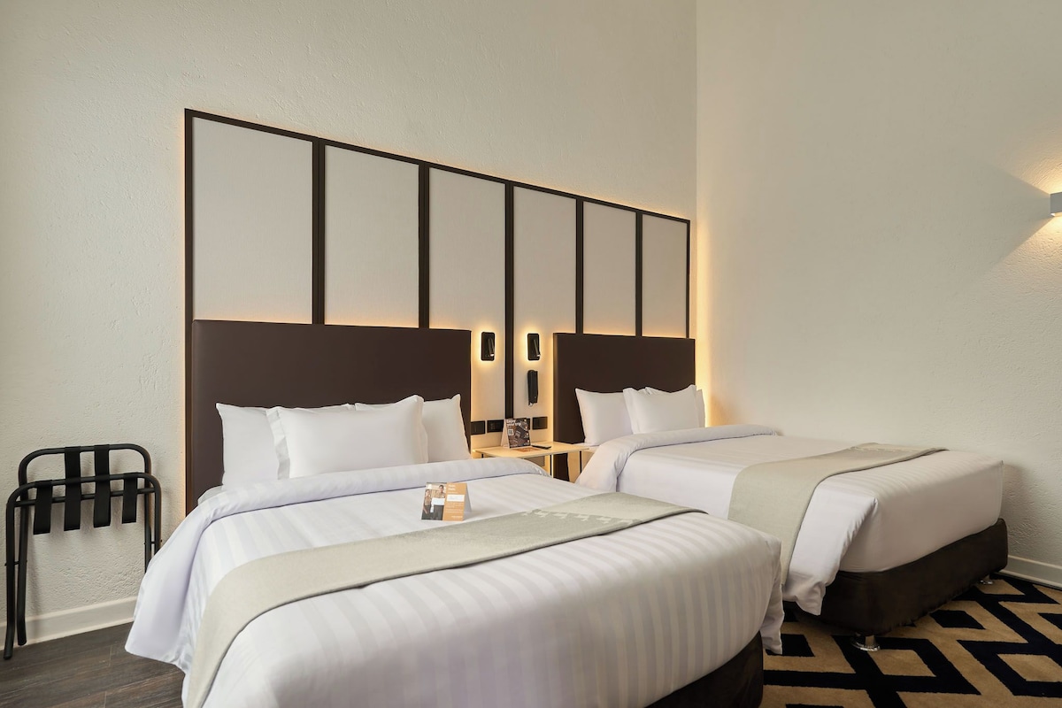 Quarto de hotel com duas camas, mesa de cabeceira e luminária na parede.
