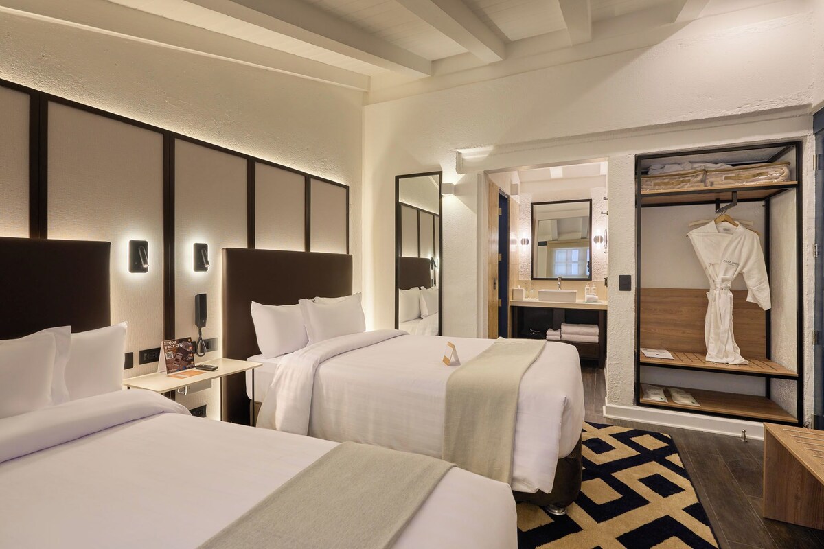 Quarto de hotel com duas camas, mesa de cabeceira com telefone e cartão, espelho, toucador com pia e roupão pendurado em um suporte.