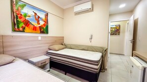 Quarto de hotel pequeno com uma cama, uma mesinha de cabeceira, ar condicionado na parede e um quadro.