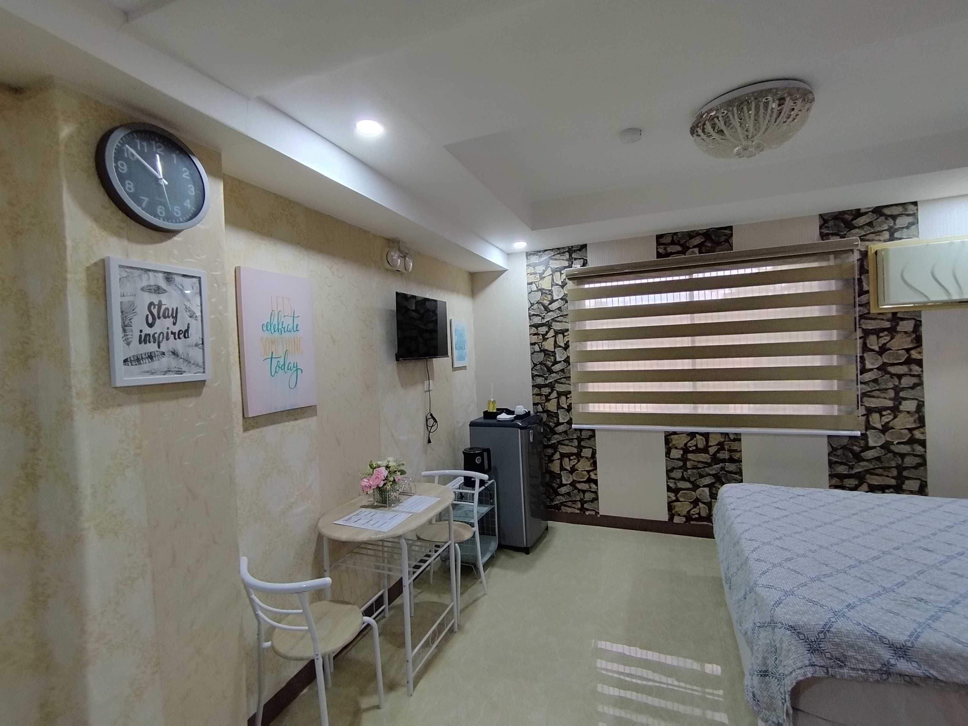 Quarto com cama, escrivaninha, cadeiras, televisão e relógio na parede.