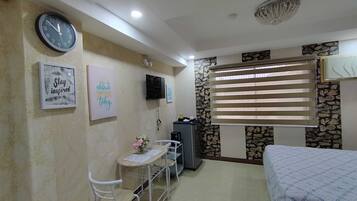 Quarto com cama, escrivaninha, cadeiras, televisão e relógio na parede.