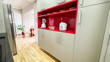 Uma cozinha com geladeira, armários, uma prateleira vermelha com eletrodomésticos e um espaço de jantar com mesa e cadeiras.