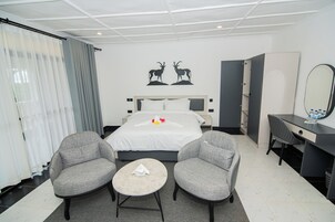 Quarto de hotel com cama, duas poltronas, uma mesa pequena, escrivaninha com espelho e guarda-roupa.