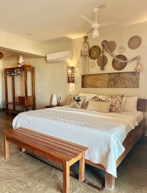 Quarto com cama, um banquinho de madeira, ventilador de teto, decoração na parede e um aparelho de ar condicionado.