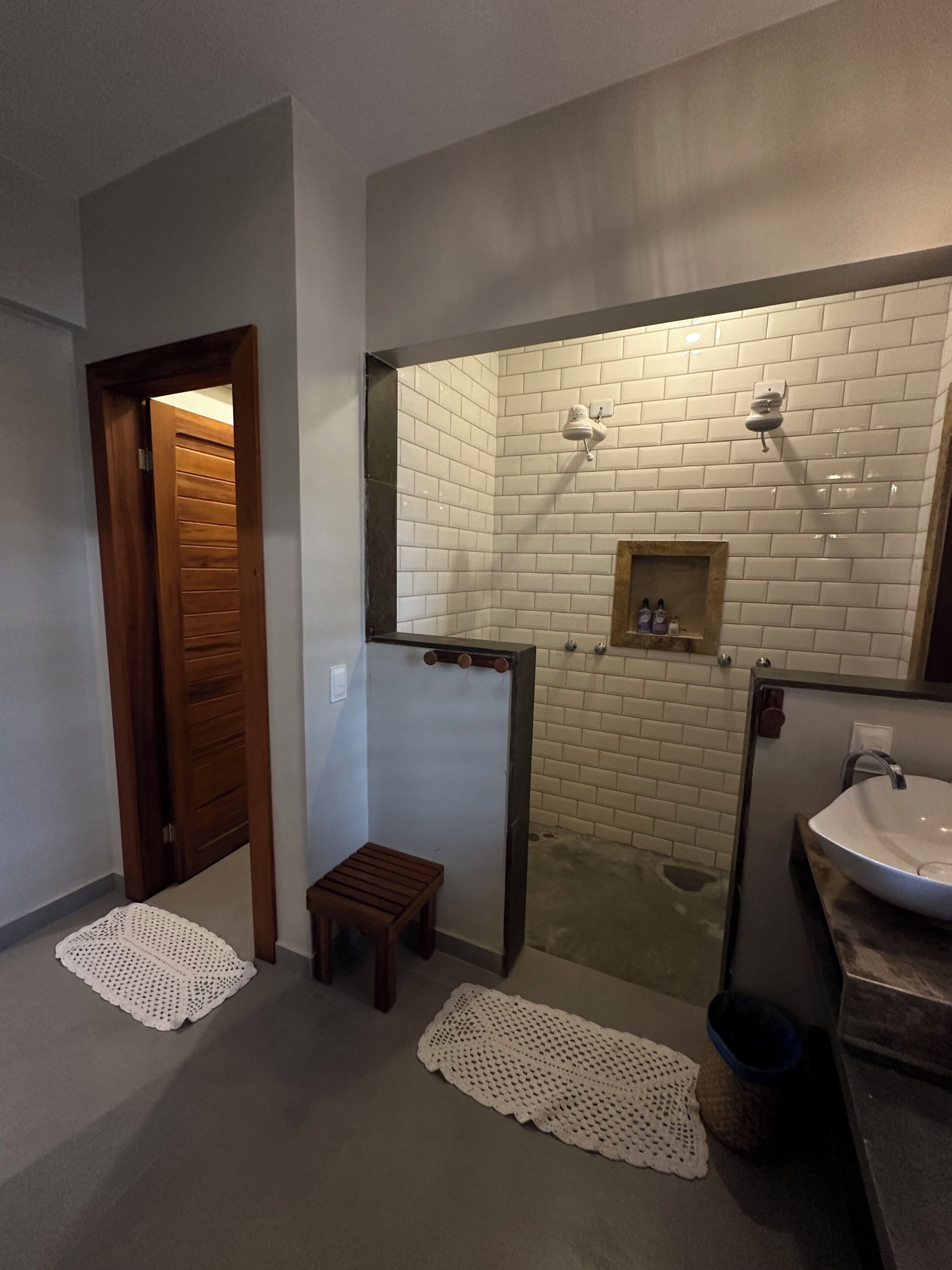 A bathroom with a white-tiled shower, a wooden door, a small wooden stool, and a sink with a mirror.