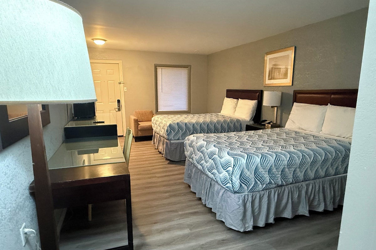 Quarto de hotel com duas camas, uma escrivaninha, uma cadeira, uma TV e uma janela com persianas.