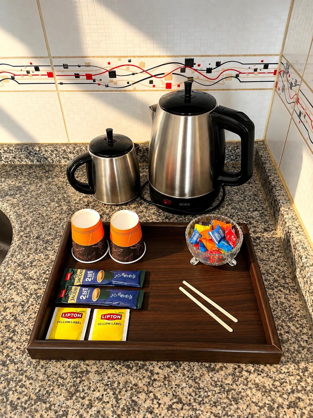 Uma bandeja com xícaras de café, chaleira, sachês de chá e uma tigela de lanches em uma bancada de cozinha.