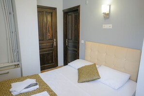 Quarto de hotel com uma cama, duas portas, uma luminária na parede e uma mesinha de cabeceira.