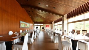 Sala de conferência com mesas e cadeiras compridas, teto de madeira e janelas amplas.