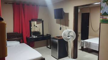 Quarto de hotel com cama, escrivaninha com computador, televisão na parede e ventilador.