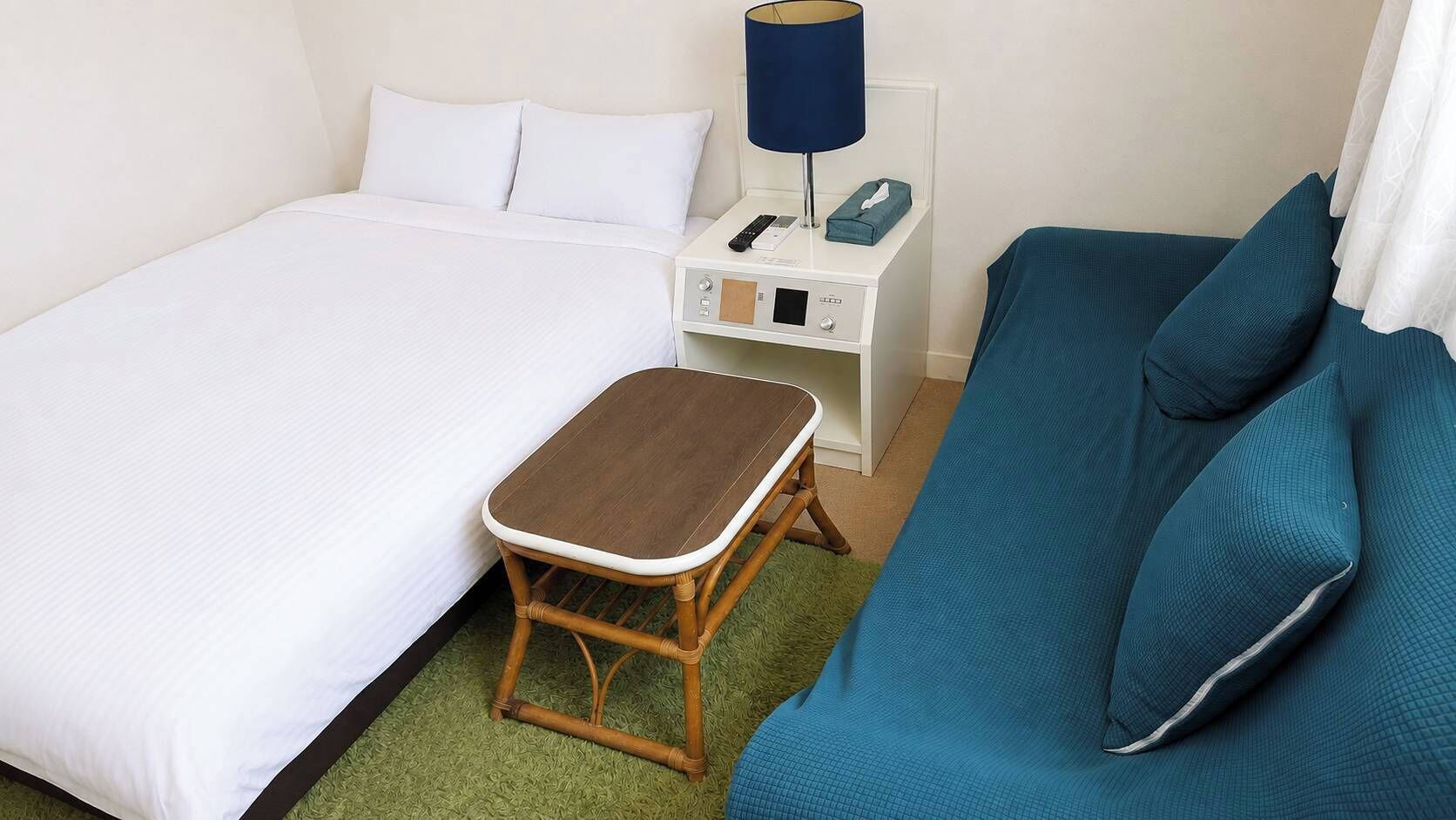 Quarto de hotel com cama, um banquinho de madeira, mesa de cabeceira com abajur e um sofá verde-água.