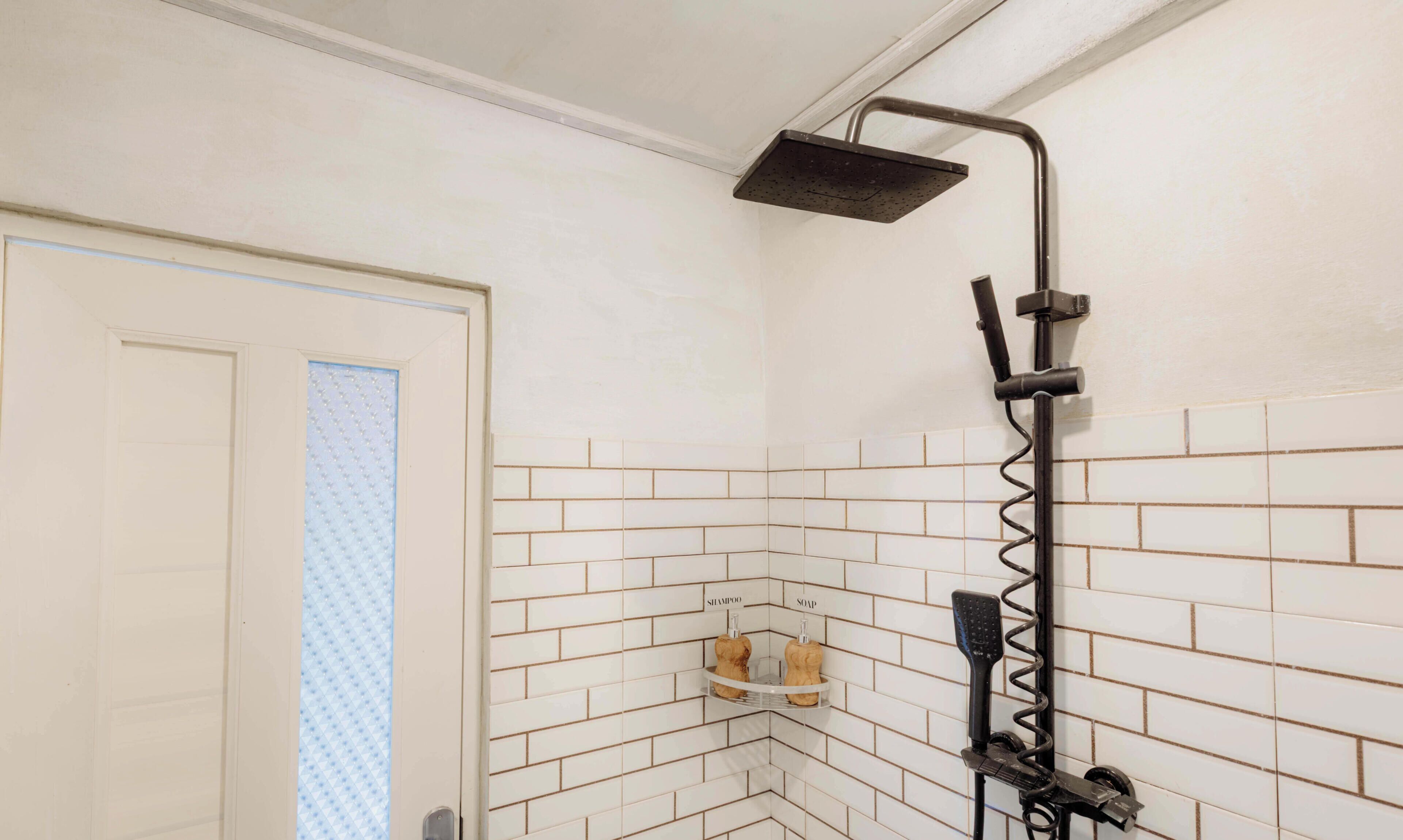 Um chuveiro no canto com um chuveiro manual, uma torneira fixada na parede e uma prateleira com garrafas de shampoo e sabonete.