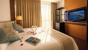 Quarto de hotel com cama, televisão, escrivaninha e janela com cortinas.