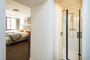 Quarto de hotel com cama, mesa de cabeceira e área de chuveiro com porta de vidro.