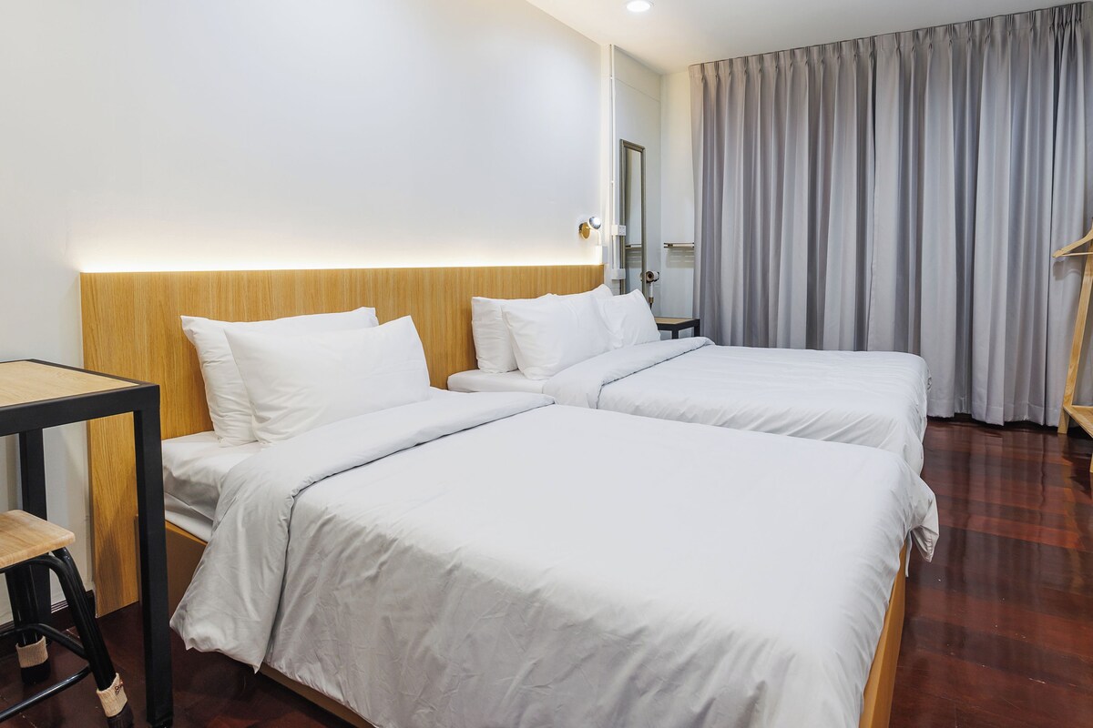 Quarto de hotel com duas camas, cabeceira de madeira, mesa de cabeceira, espelho, cabideiro de madeira e janela com cortinas.