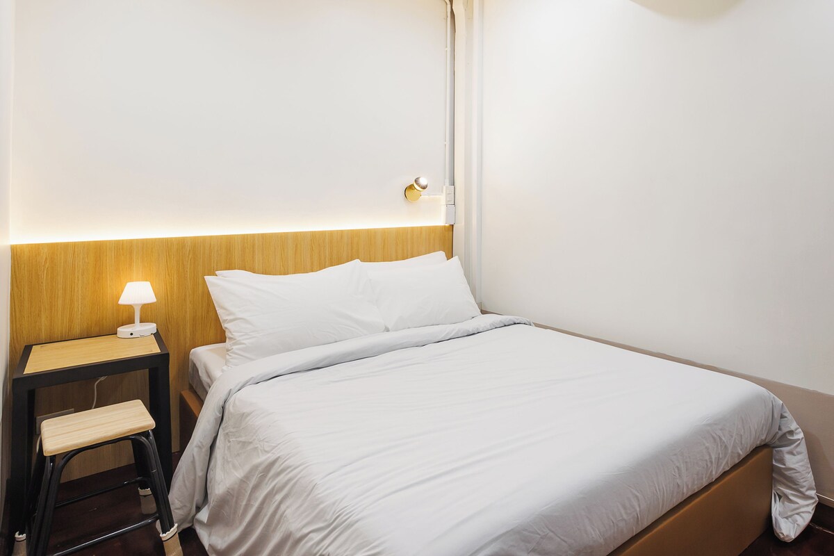 Cama bem arrumada com roupa de cama branca, mesa de cabeceira com abajur e cabeceira de madeira.