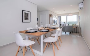 Uma zona de refeições moderna com uma mesa de madeira posta para seis pessoas, cadeiras brancas e uma obra de arte emoldurada na parede.