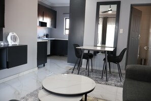 Uma sala moderna com uma mesa redonda, duas cadeiras pretas, um sofá cinzento, uma cozinha com mesa de jantar e um ventilador de teto.
