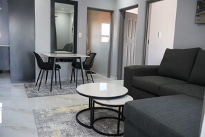 Uma sala de estar moderna com um sofá cinzento, uma mesa de centro redonda e uma área de jantar com uma mesa branca e cadeiras pretas.