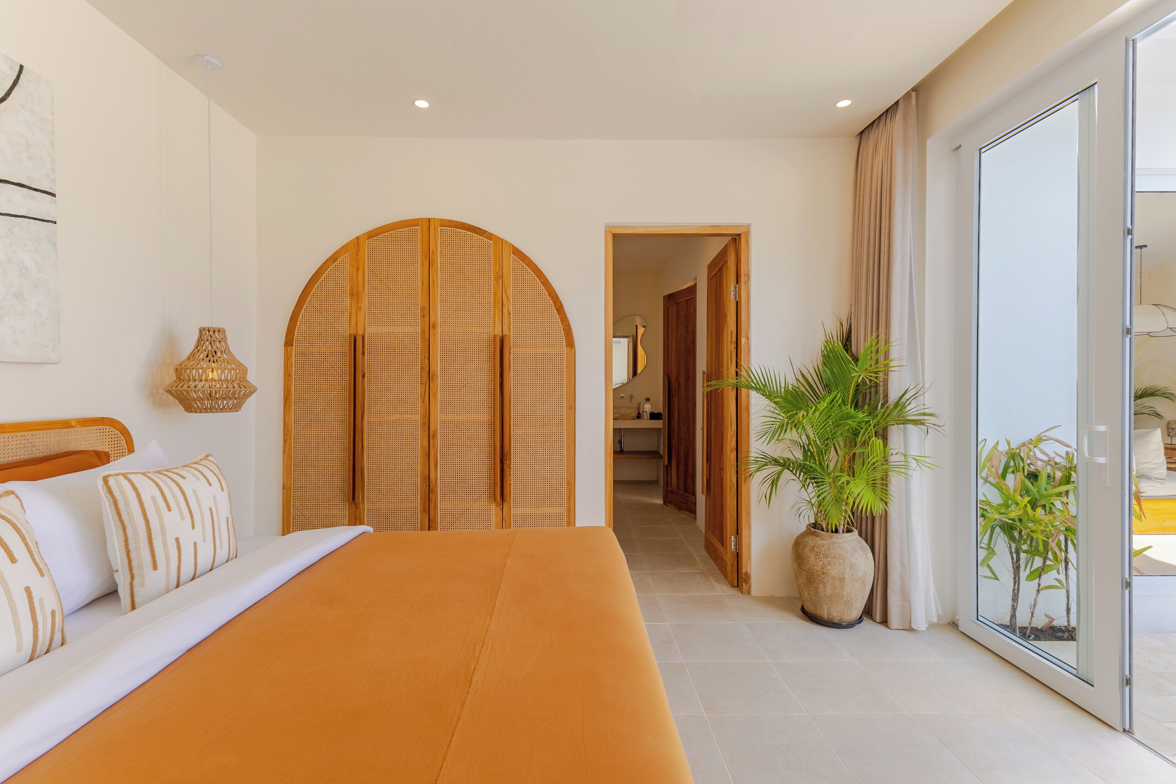 Quarto de hotel moderno com uma cama grande, um guarda-roupa de madeira, uma planta em vaso e uma porta de vidro deslizante para a varanda.