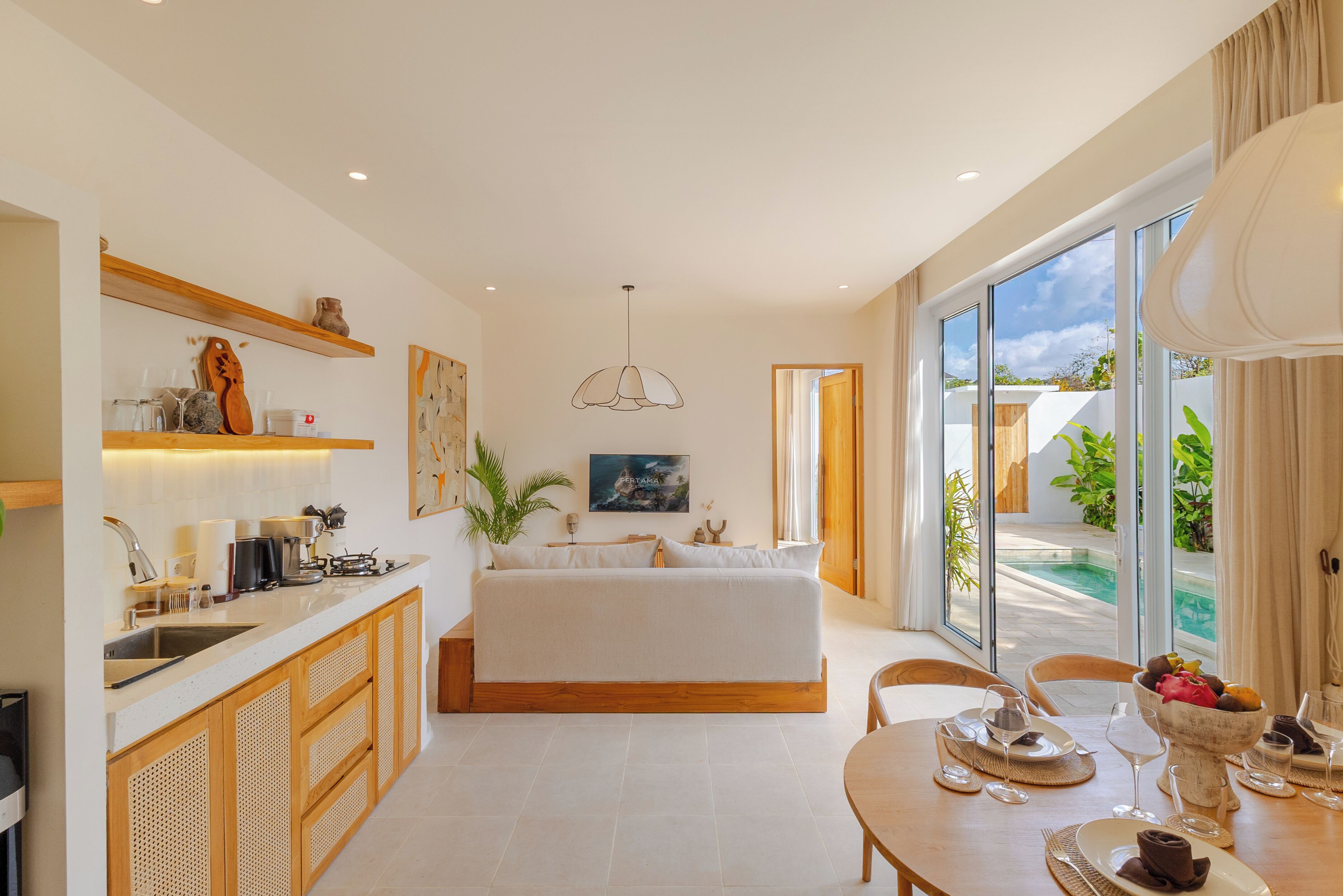 Quarto de hotel moderno com uma cama grande, área de jantar com mesa posta para dois e vista para uma piscina.