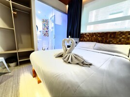 Quarto de hotel com cama bem arrumada, guarda-roupa, banheiro visível através de uma porta de vidro e parede com padrão de tijolos.