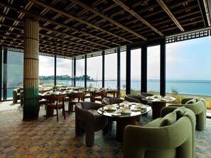 Um restaurante com mesas e cadeiras redondas, janelas amplas e vista para o mar.