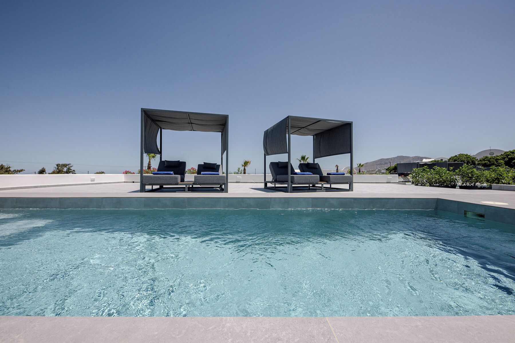 Piscina com duas espreguiçadeiras sob um toldo, céu claro e colinas ao longe.