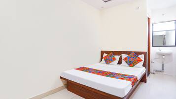 Quarto com estrutura de cama de madeira, roupa de cama branca e almofadas geométricas coloridas.
