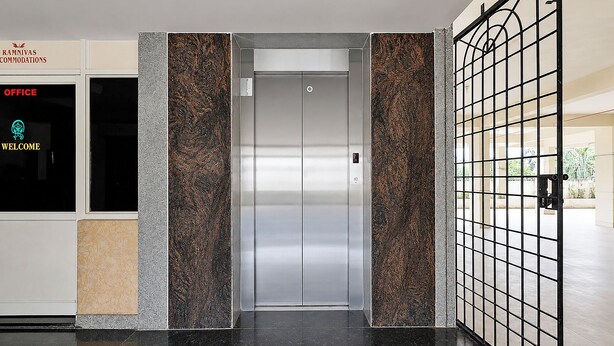Um elevador com acabamento metálico, ladeado por paredes texturizadas e um portão de metal.
