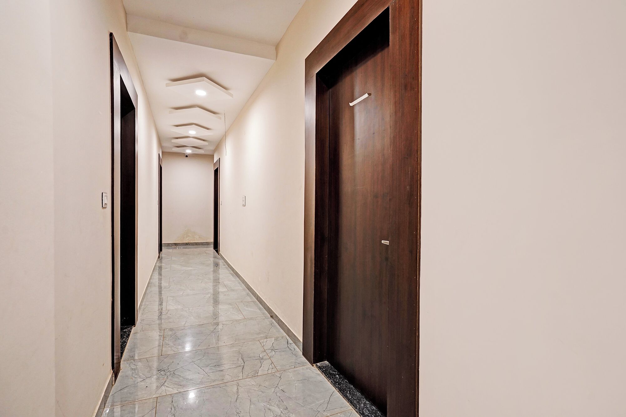 Um corredor com piso de mármore, porta de madeira e teto com iluminação embutida.