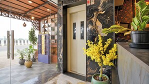 Uma área de recepção de hotel com parede de mármore, balcão de recepção, planta em vaso e uma porta com placa de número.