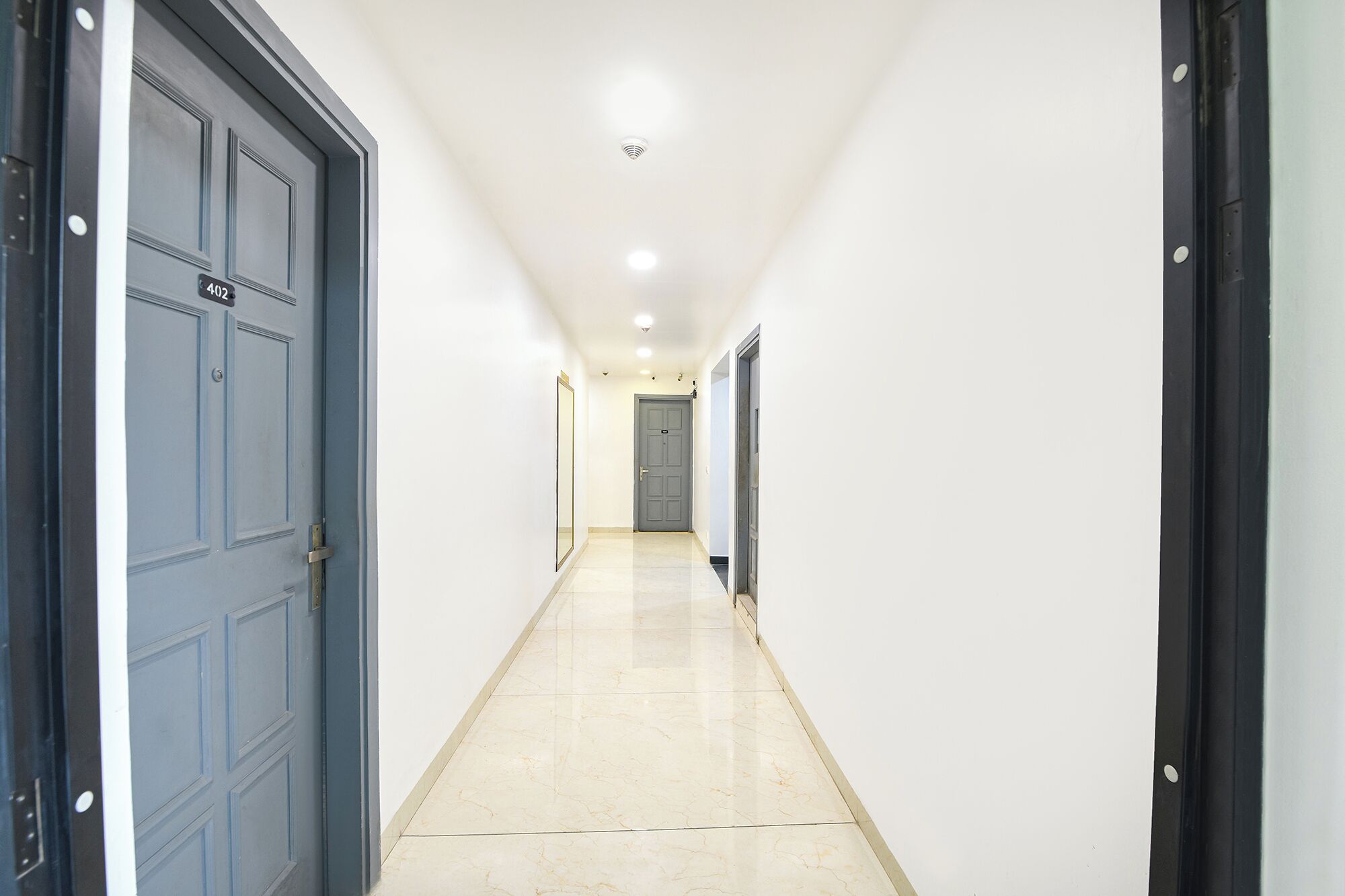 Um corredor com uma porta marcada '402' à esquerda, um espelho à direita e uma porta no final.
