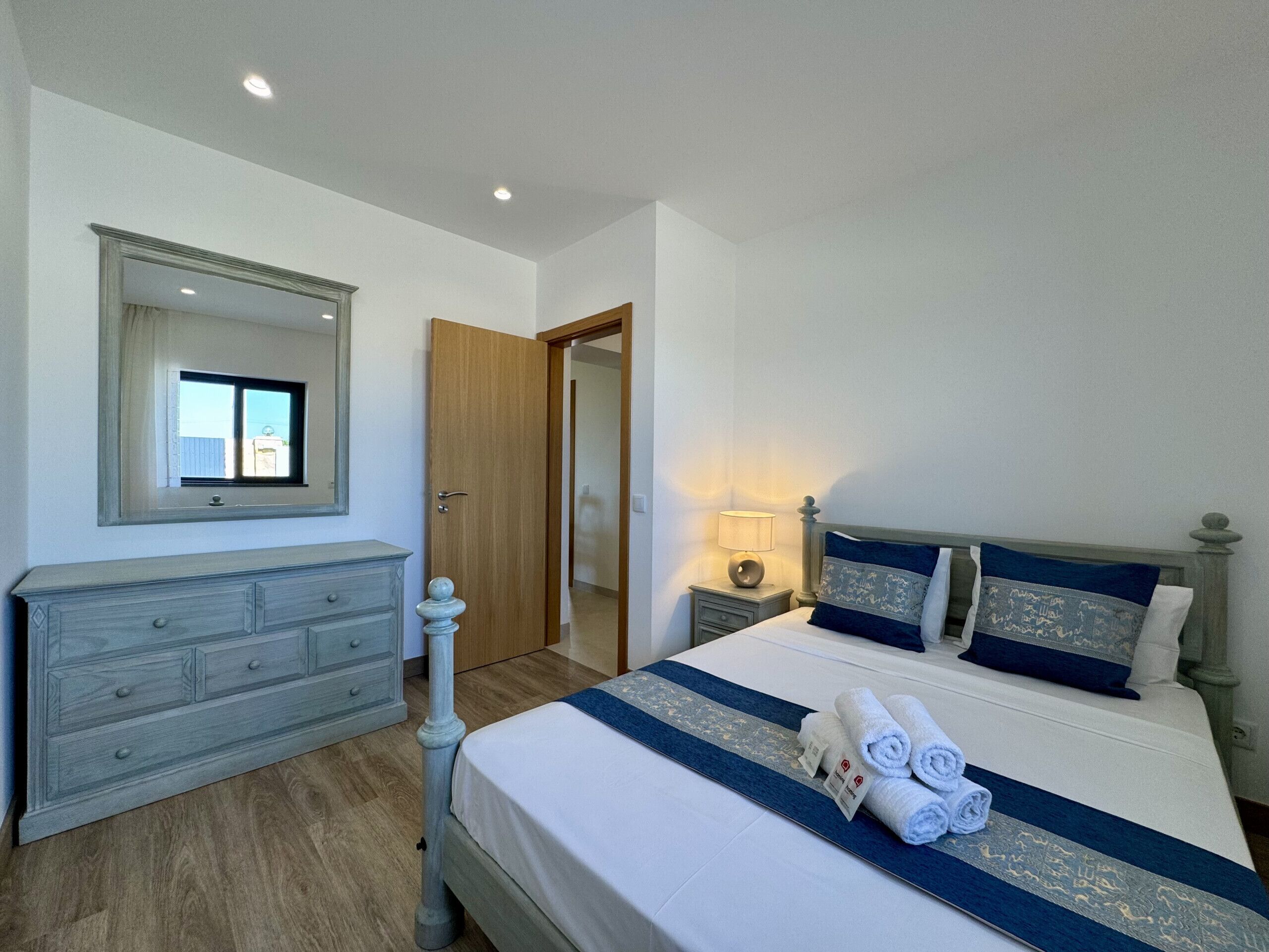 Quarto de hotel com cama, penteadeira, espelho e mesa de cabeceira com abajur.