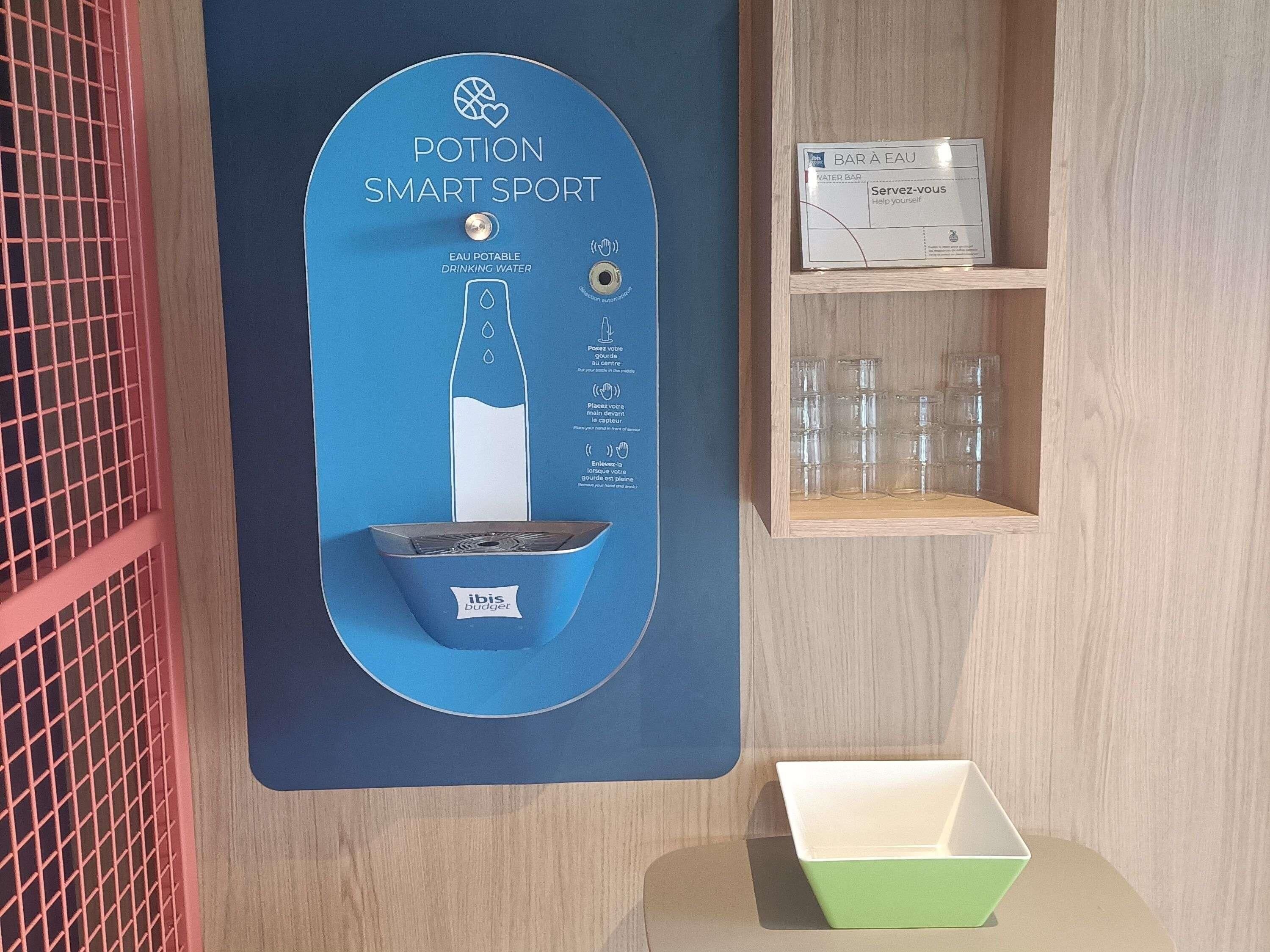 Um dispensador fixado na parede com uma garrafa e uma tigela, rotulado 'Potion Smart Sport', e um bar de água com copos.