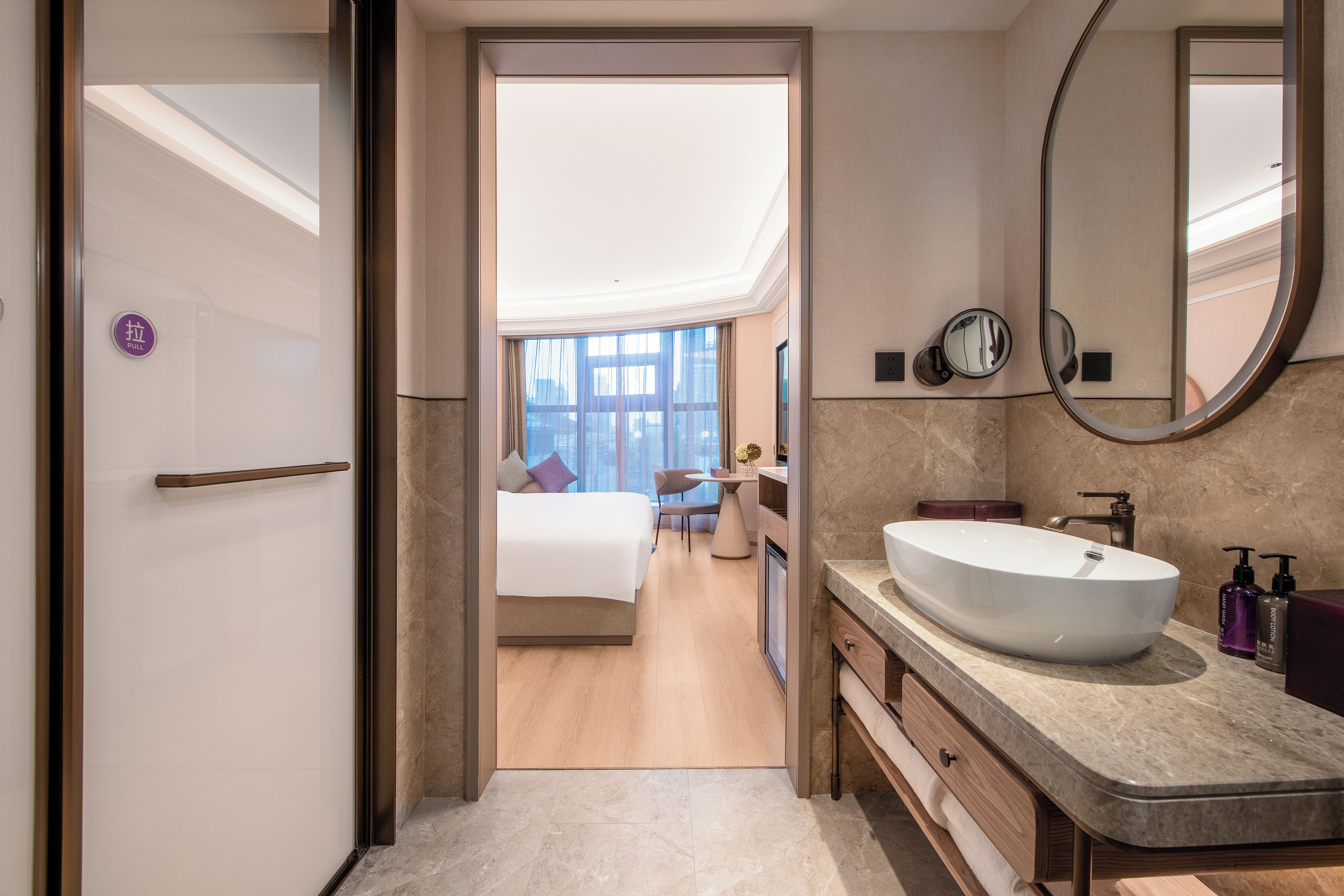 Quarto de hotel com uma cama grande, banheiro com pia e espelho amplos e vista da cidade pela janela.