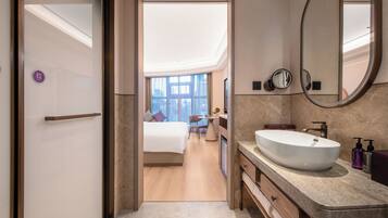 Quarto de hotel com uma cama grande, banheiro com pia e espelho amplos e vista da cidade pela janela.